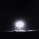 Black-and-white photo of a pitch-black night sky with brilliant white glowing nuclear cloud in distance at center.