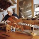 photo of man with white hair sitting behind desk and holding acoustic guitar and surrounded by other musical instruments, with large antique portrait of man playing violin in background