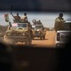 photo through car window of military vehicles and trucks driving on dirt road, one flying flag, with fighters, arms raised, sitting on tops and sides