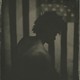 A black man is silhouetted by an American flag in a black and white tintype photo